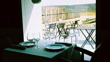 Mesa de jantar posta com pratos e taças de vinho, com vista para uma paisagem cênica de turbinas eólicas e colinas.