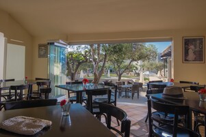 Uma área de refeições ao ar livre com mesas e cadeiras, vista para árvores e um edifício ao fundo, e um quadro na parede.