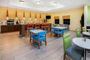 Uma sala de jantar de hotel com várias mesas e cadeiras, uma área de cozinha e uma televisão na parede.