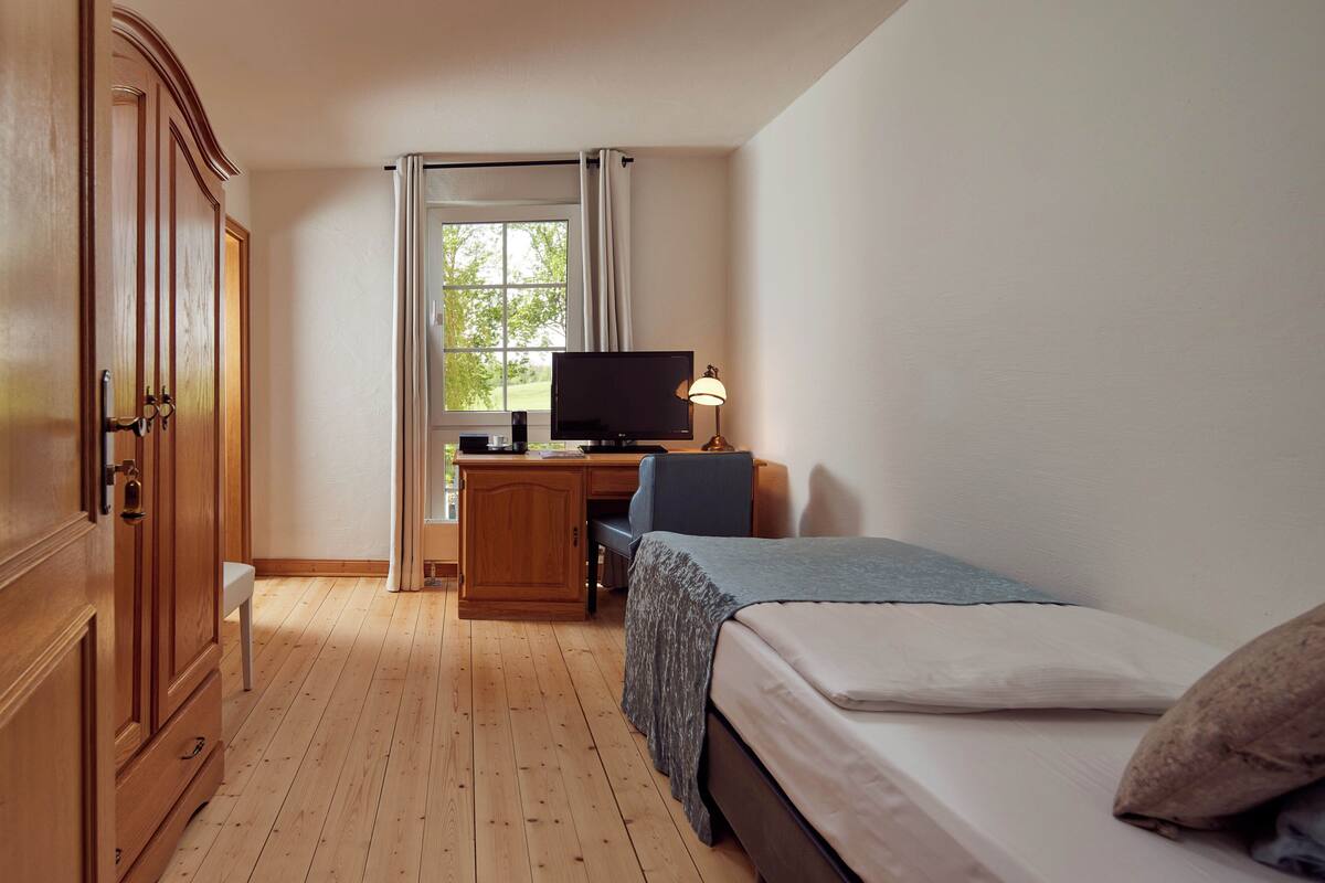Quarto de hotel com guarda-roupa de madeira, uma mesa com computador e televisão, cama com manta, e janela com cortinas.