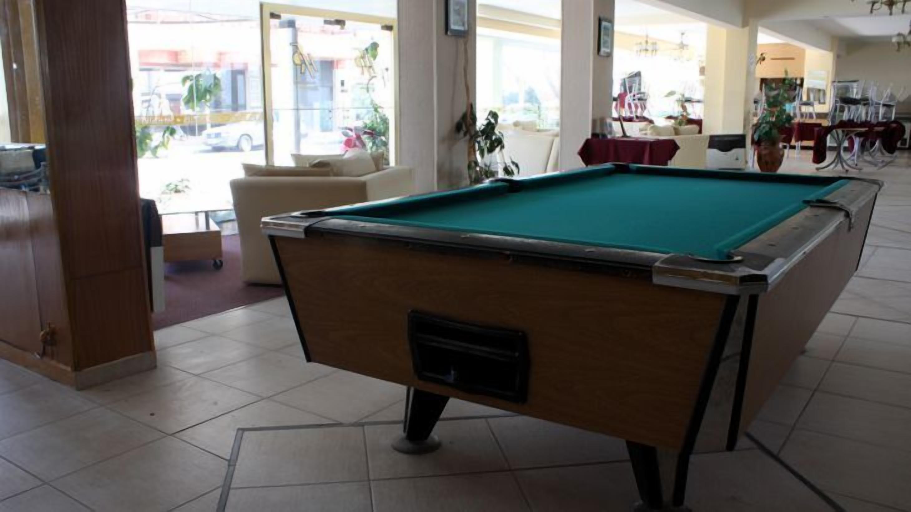 Uma mesa de bilhar em um salão com piso ladrilhado, janelas amplas e vista para a rua.