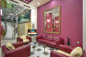 Ampla sala de estar com sofá vermelho, poltronas bege e uma mesa de centro de vidro. Um espelho grande e um quadro decoram as paredes, e uma escada é visível ao fundo.