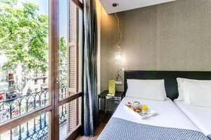 Quarto de hotel com uma janela grande, uma cama com lençóis brancos, uma mesinha de cabeceira com abajur e uma bandeja com itens de café da manhã.