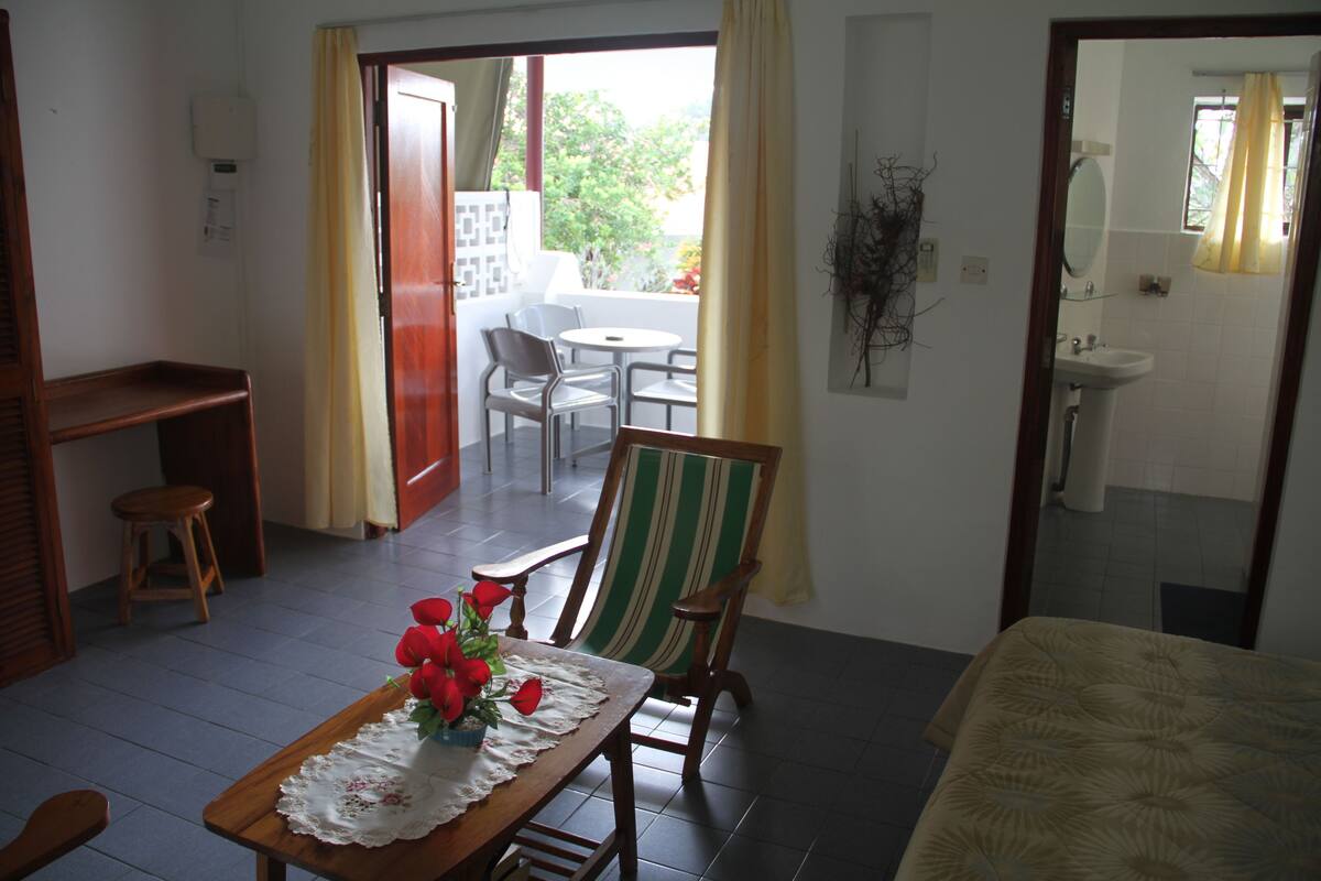 Quarto com mesa de jantar posta com toalha rendada e flores vermelhas, uma cadeira de madeira, uma cama, banheiro com pia e espelho, e vista para o exterior por uma porta.