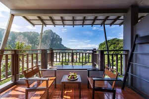 Varanda com mobiliário de madeira, uma mesa com frutas e vista para montanhas e vegetação.
