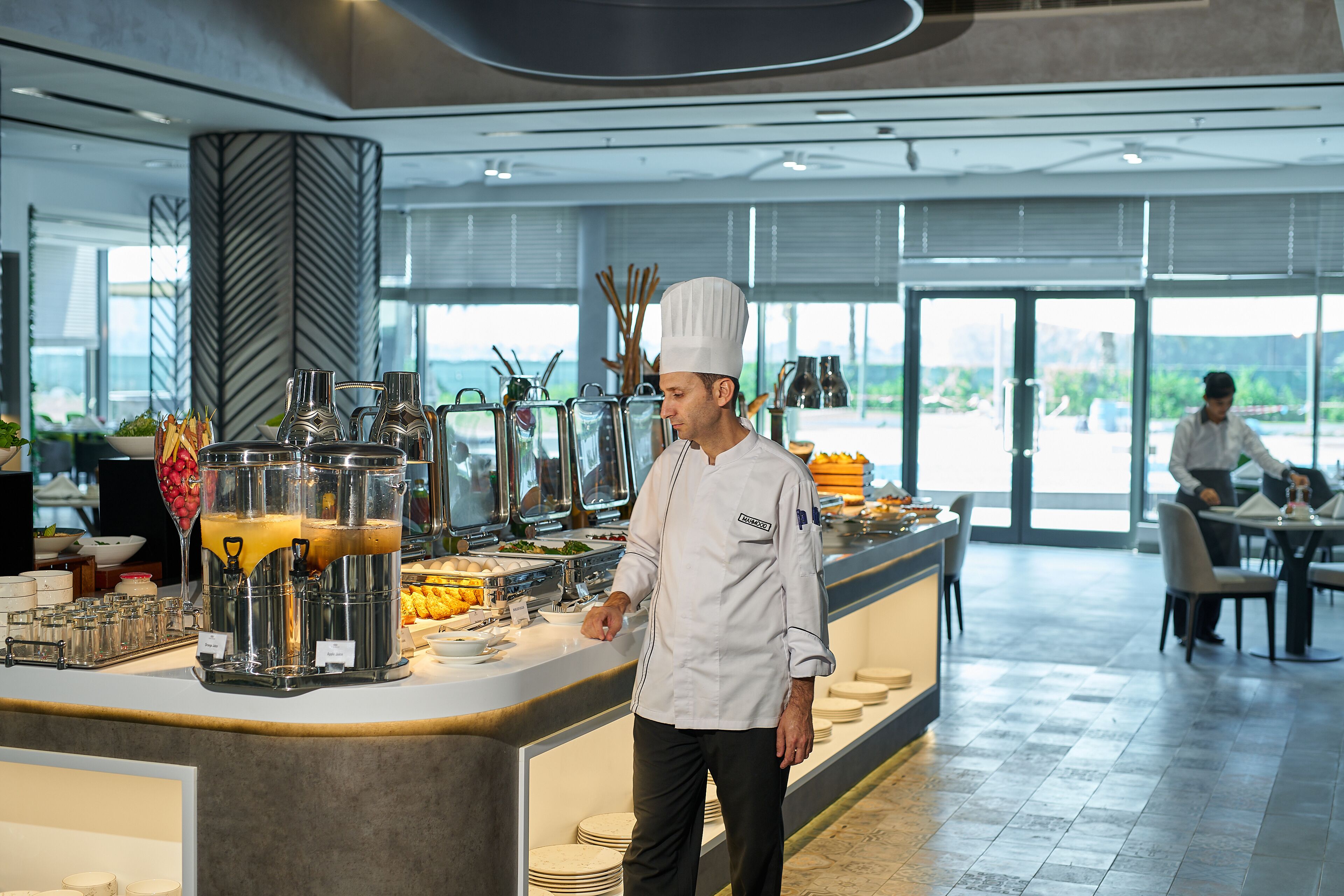 Restaurante moderno com serviço de buffet, incluindo diversos recipientes de comida e um chefe de cozinha com uniforme.