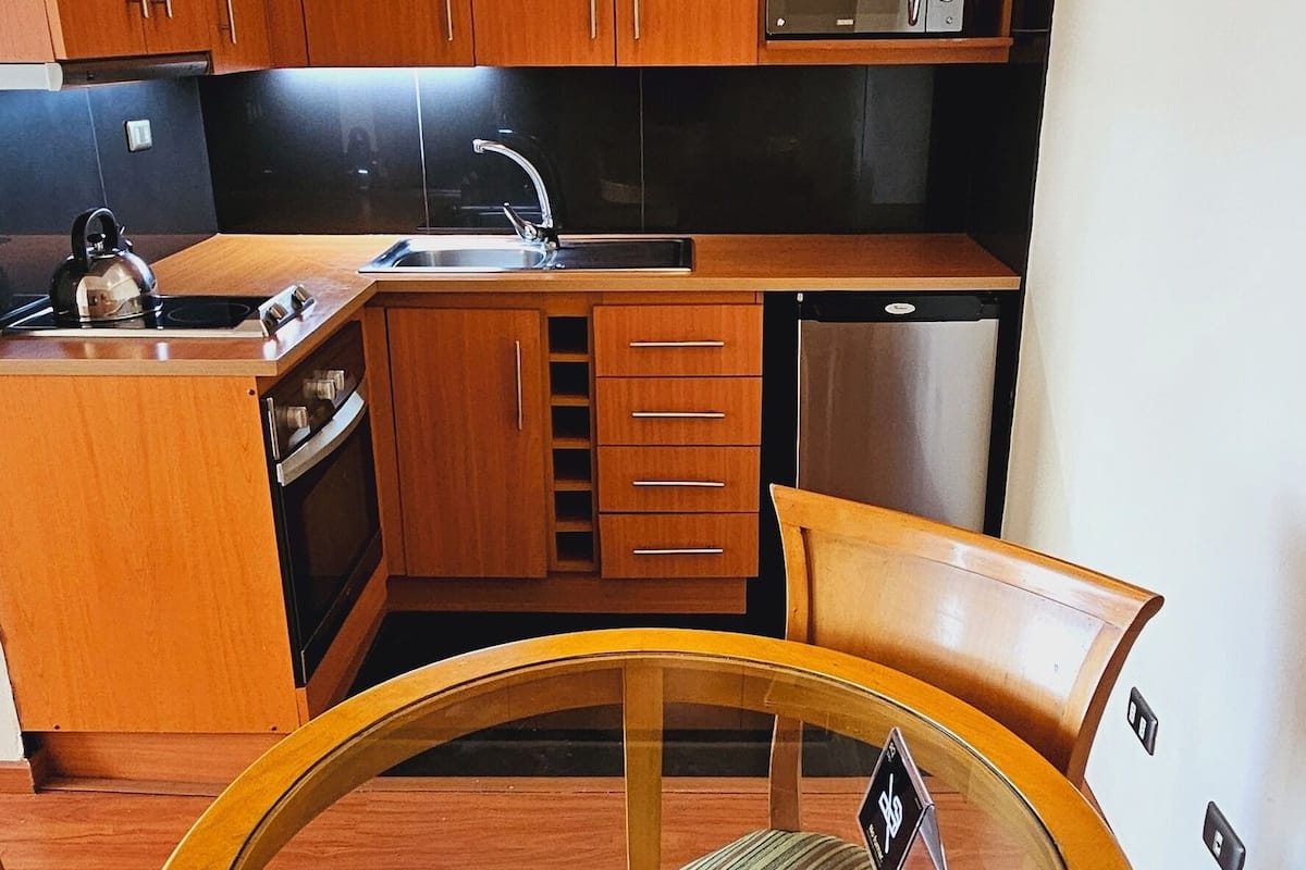 Cozinha compacta com armários de madeira, micro-ondas, pia e um refrigerador pequeno. Há uma mesa redonda de madeira com duas cadeiras e tampo de vidro.