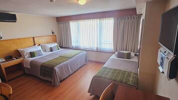Quarto de hotel com duas camas, televisão, escrivaninha com telefone e janela com cortinas.
