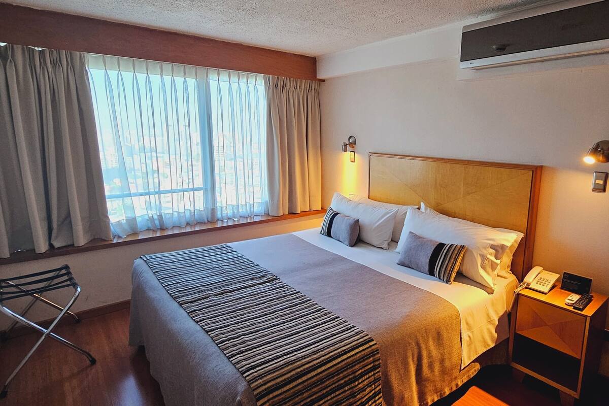 Quarto de hotel com cama, televisão fixada na parede, mesa de cabeceira com telefone e janela com cortinas.