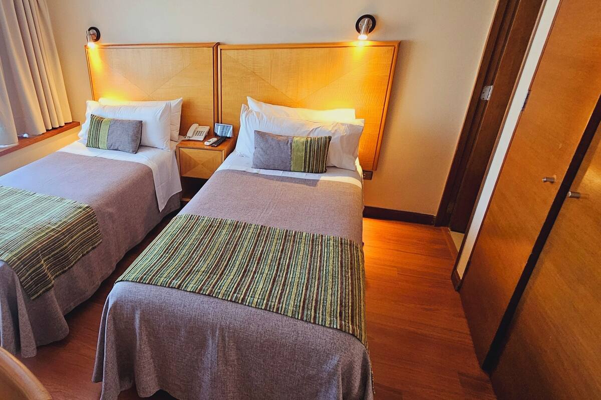 Quarto de hotel com duas camas de solteiro, cabeceira de madeira, mesa de cabeceira com abajur e uma janela com cortinas.