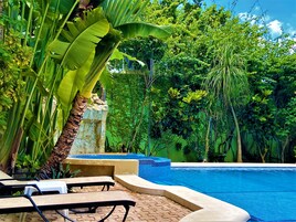 Uma área de piscina com espreguiçadeiras, hidromassagem e vegetação tropical exuberante.