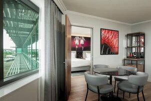 Quarto de hotel moderno com uma janela grande, uma cama, uma mesa redonda com cadeiras e uma estante de livros.