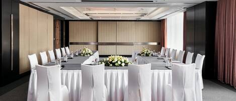 Uma sala de conferências com uma mesa comprida, cadeiras brancas e centros de mesa com flores.