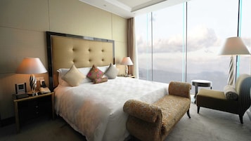 Quarto de hotel com uma cama grande, duas mesas de cabeceira, uma cadeira, uma pequena mesa e vista da cidade através de grandes janelas.