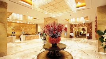 Um saguão de hotel com balcão de recepção, um vaso decorativo com folhagem vermelha, um candelabro e piso de mármore.