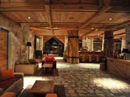 Um hall amplo com piso de pedra, vigas de madeira no teto e uma coluna de madeira grande. Há um espaço de bar com banquetas e balcão.