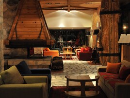 Uma sala de estar acolhedora com lareira de pedra, vigas de madeira e diversas opções de assento, incluindo um sofá e poltronas.