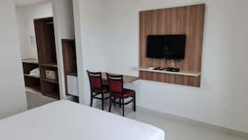 Quarto de hotel com uma cama, uma mesa com duas cadeiras, uma TV fixada na parede e uma pequena prateleira com um controle remoto.