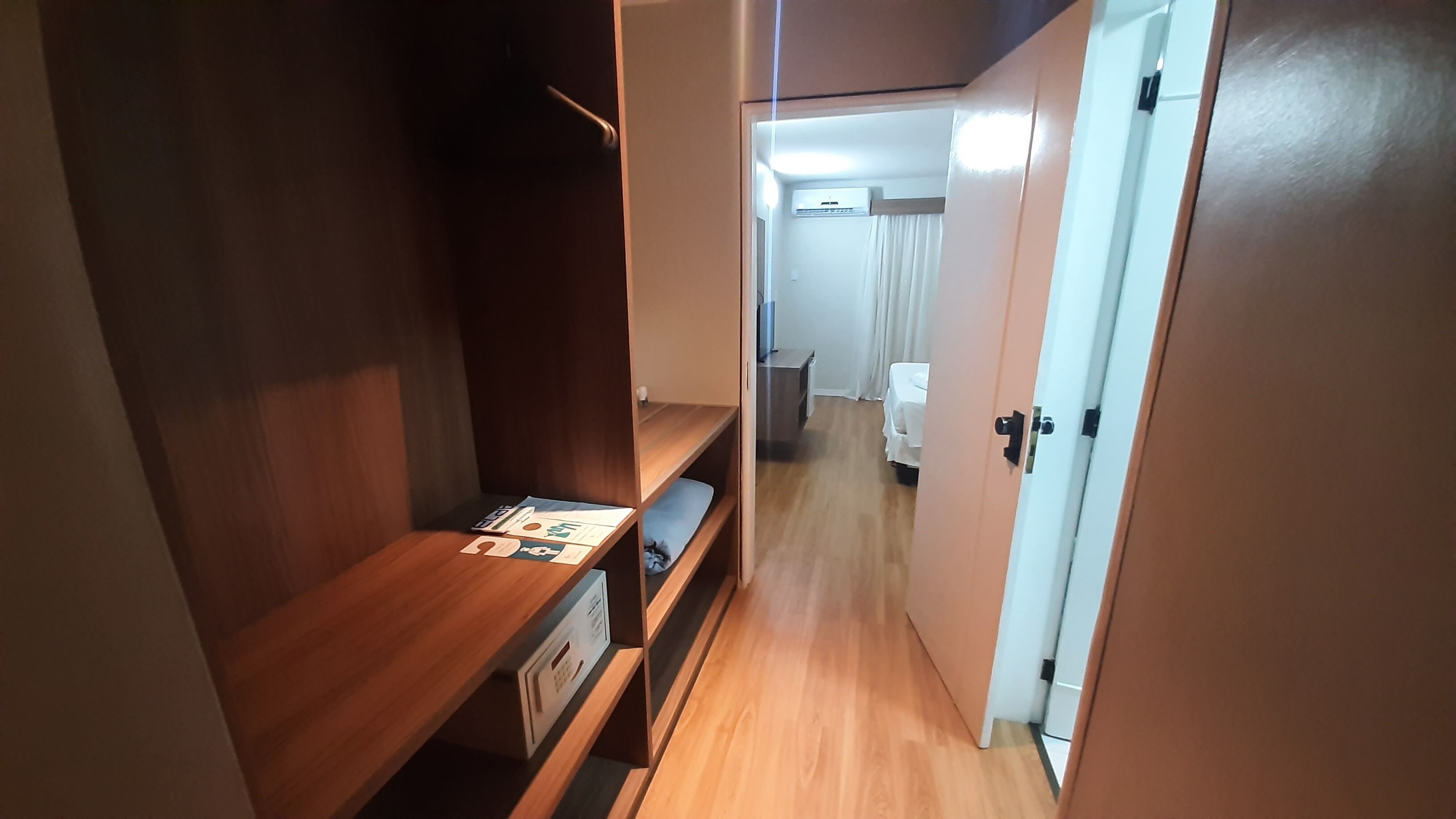 Um corredor estreito com piso de madeira, um banco de madeira com papéis e uma porta que dá para um quarto com cama e cortinas.