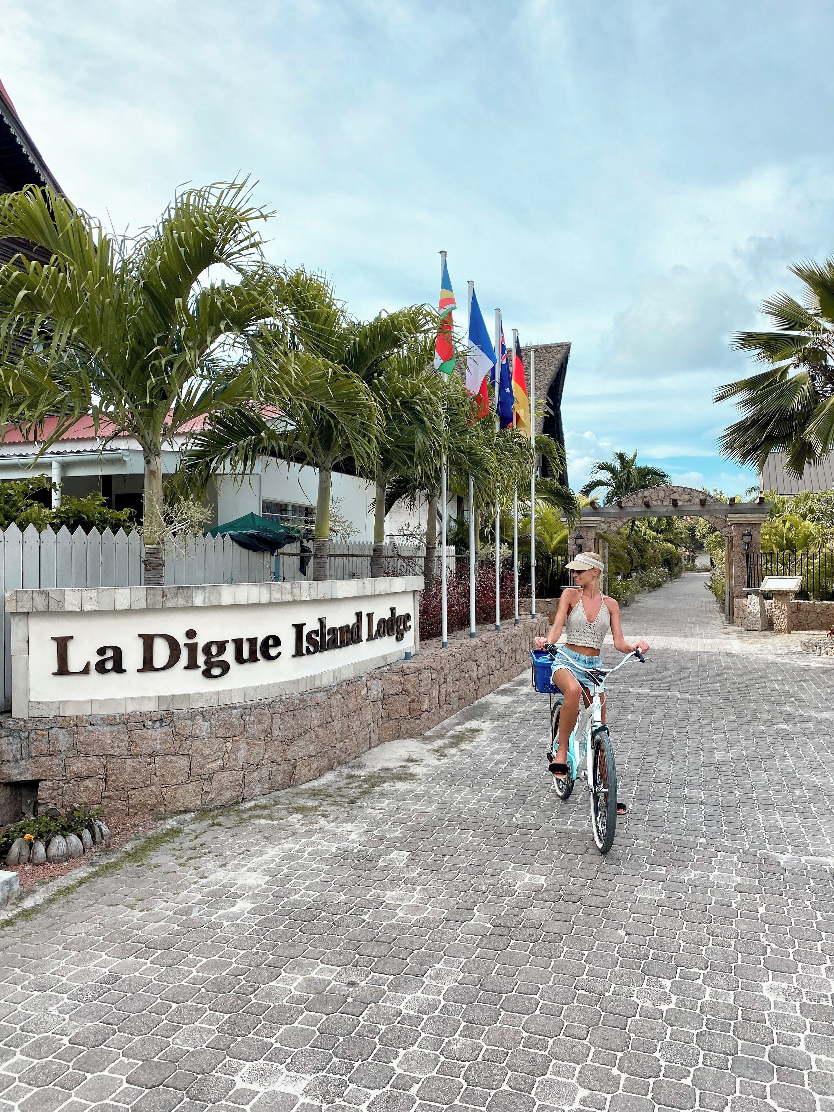 Placa para a Pousada Ilha de La Digue, com um caminho pavimentado e palmeiras.