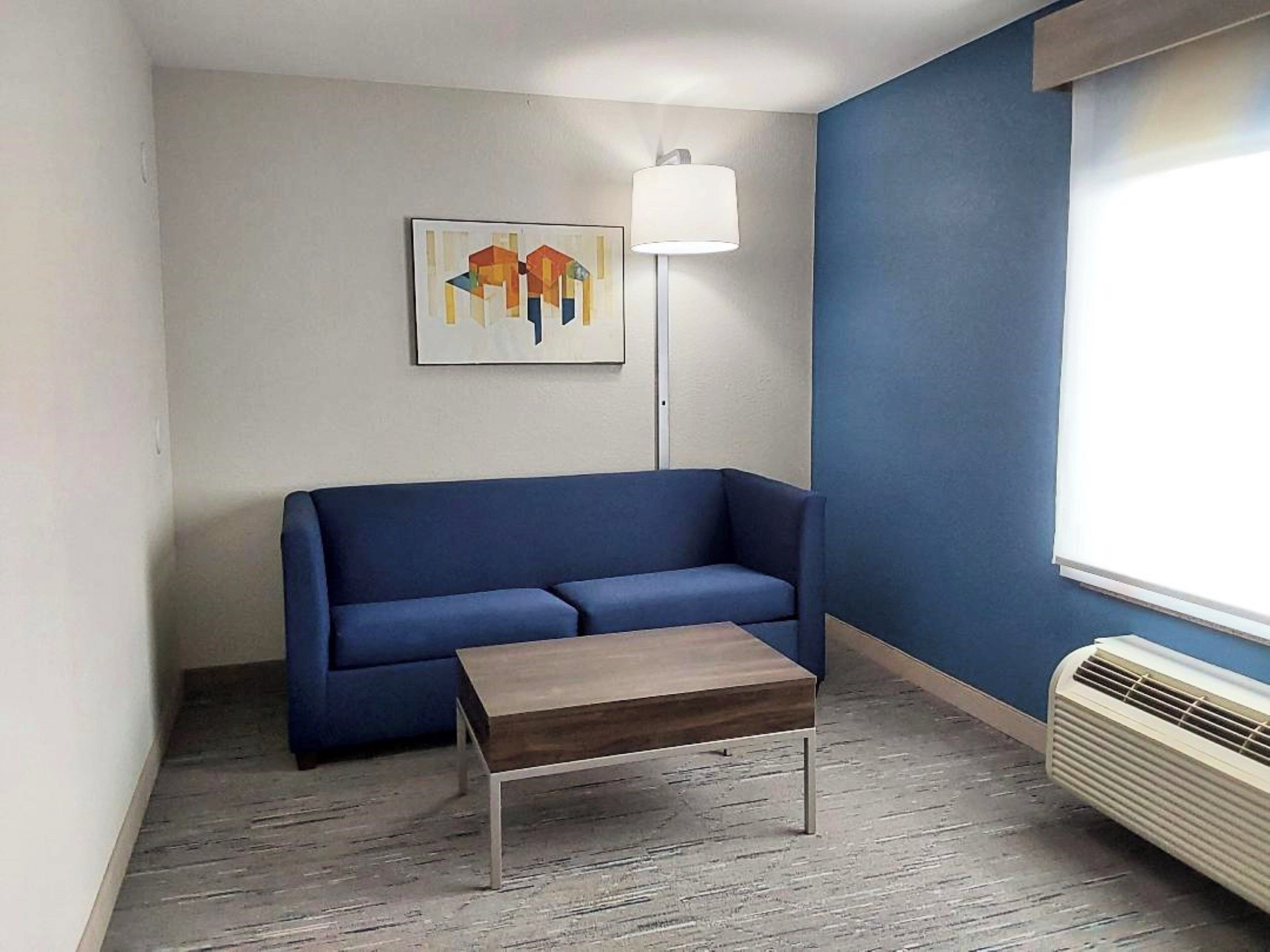 Um quarto com um sofá azul, uma mesa de centro de madeira, um abajur e uma janela com ar condicionado.