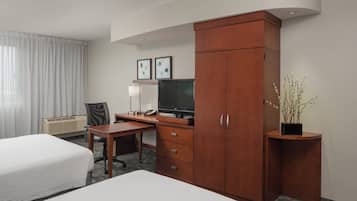 Quarto de hotel com duas camas, uma escrivaninha com televisão, um guarda-roupa e um vaso de planta.