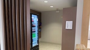 Corredor com uma máquina de vendas repleta de bebidas, um painel de madeira e uma almofada estampada.
