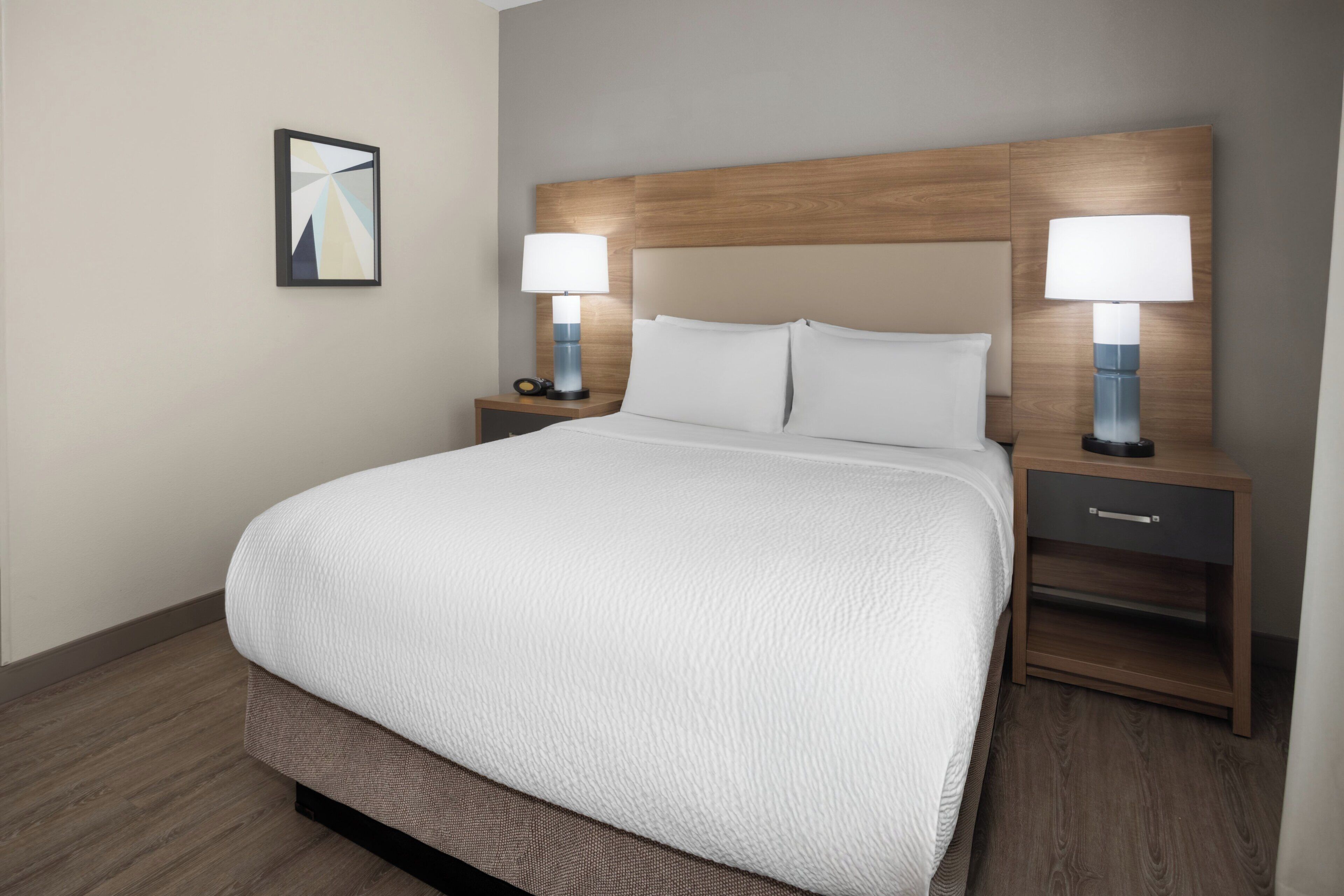 Cama bem arrumada com edredom branco e duas almofadas, com mesas de cabeceira de madeira com luminárias e uma gaveta.