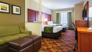 Quarto de hotel com um sofá verde, um banquinho marrom, uma cama com roupa de cama branca e um cobertor verde, uma escrivaninha com luminária e uma televisão sobre um móvel de madeira.