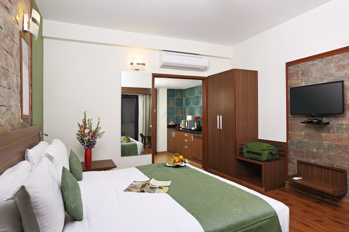 Quarto de hotel com uma cama, manta verde, tigela de frutas, bolsa verde, televisão e parede de pedra.