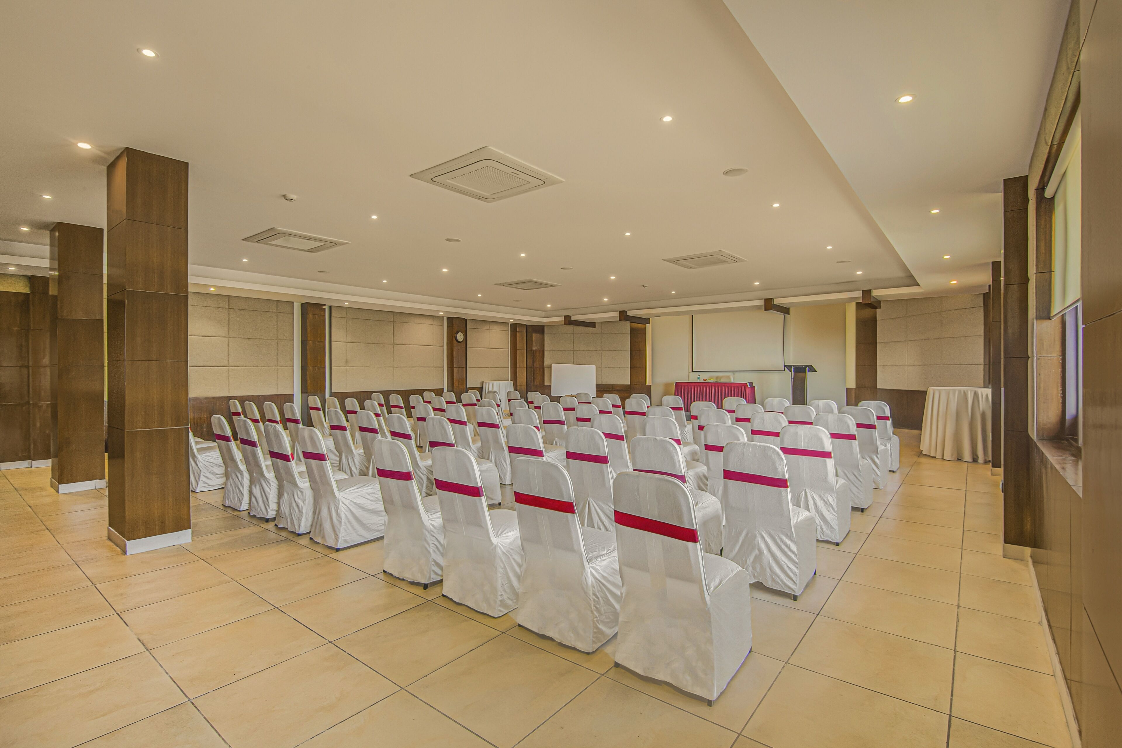 Uma grande sala de conferências com fileiras de cadeiras cobertas com tecido branco e fitas cor-de-rosa, um pódio e uma tela de projeção.