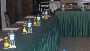 Uma sala de conferências com mesas compridas cobertas por toalhas verdes, equipada com garrafas de água, copos e blocos de anotações.