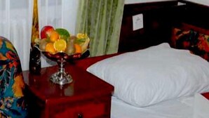 Quarto de hotel com cama, mesa de cabeceira com fruteira e uma cadeira colorida.