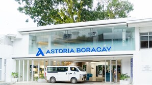 Um edifício moderno com fachada branca e grandes janelas de vidro. Uma van branca está estacionada em frente, e a placa acima diz "ASTORIA BORACAY".