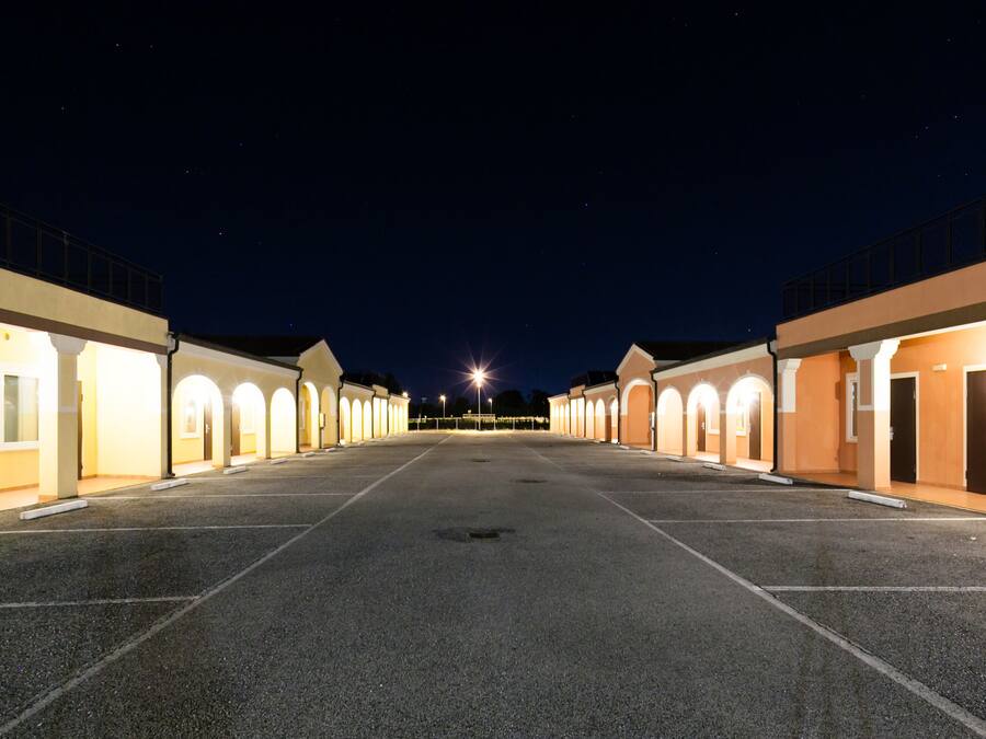 Uma fileira de quartos de motel com uma passarela central, cada quarto possui porta e uma pequena janela, e o exterior é iluminado por luminárias.