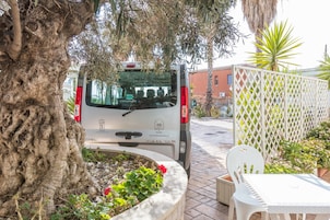 Uma van estacionada com um logotipo e texto na parte traseira, uma cadeira de plástico branca, uma mesa e um canteiro com plantas.