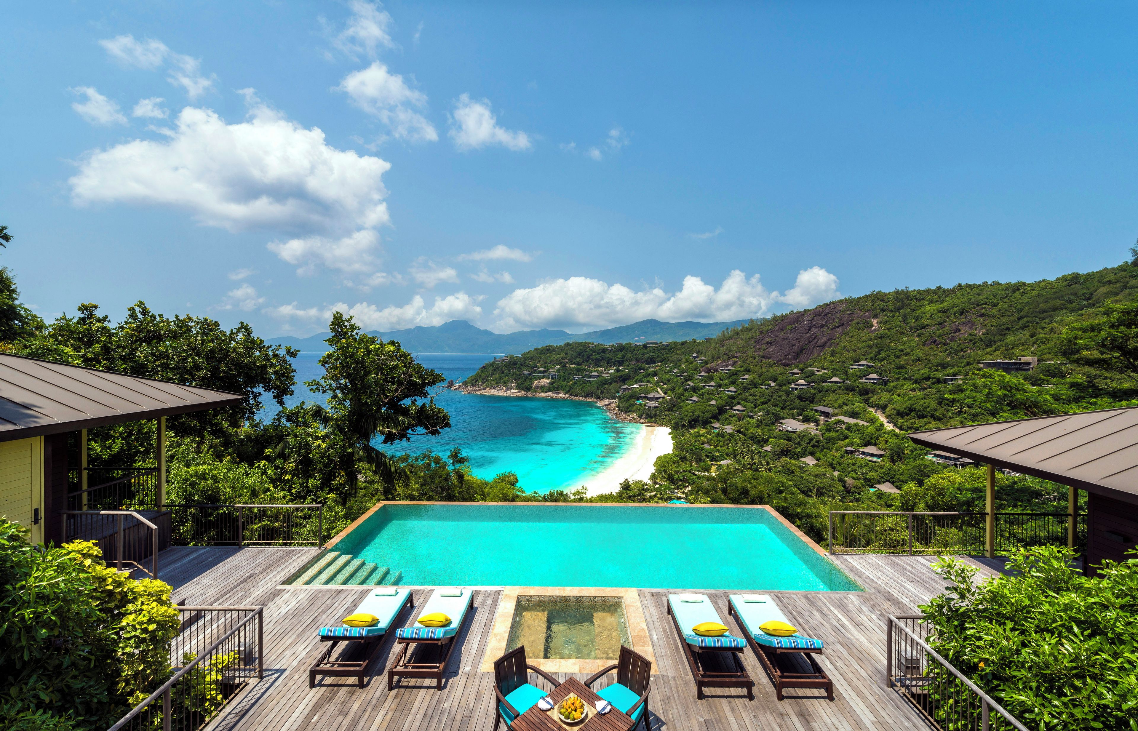 Piscina com espreguiçadeiras e mesa, com vista para uma praia e montanhas.