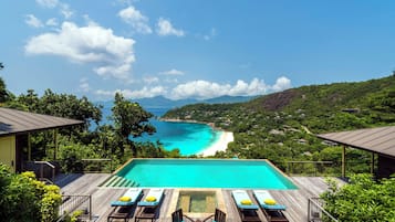 Piscina com espreguiçadeiras e mesa, com vista para uma praia e montanhas.