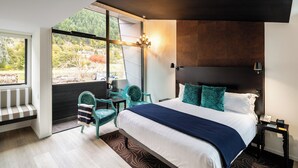 Quarto de hotel moderno com uma cama grande, vista para uma floresta, uma pequena mesa com cadeiras e um banco junto à janela.