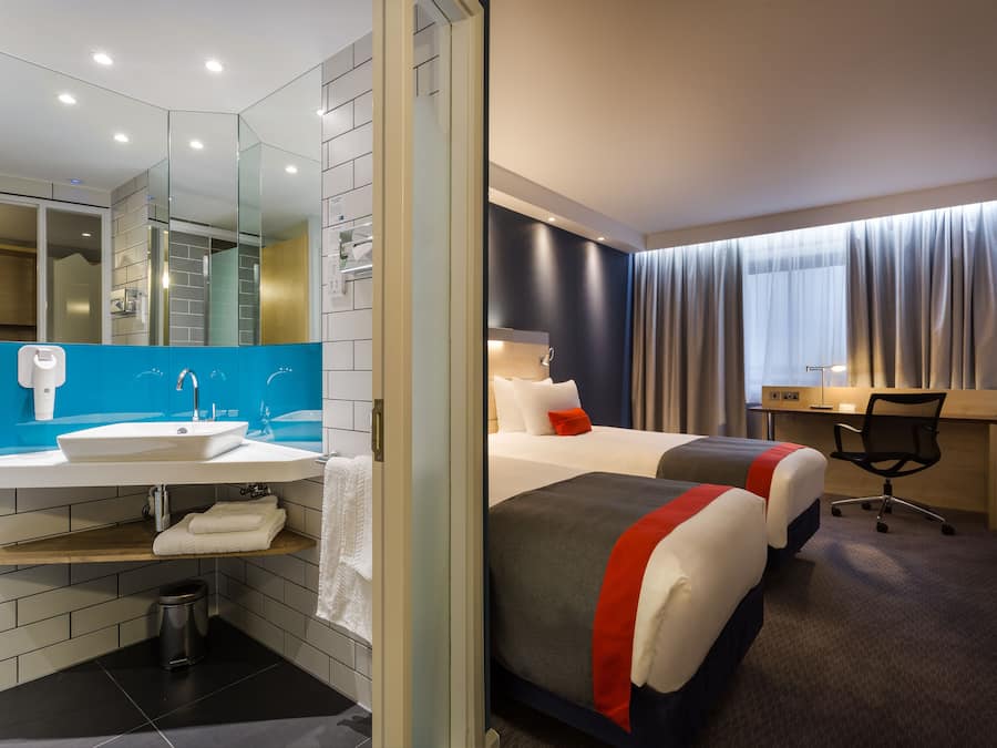 Quarto de hotel com banheiro, cama, escrivaninha e televisão.