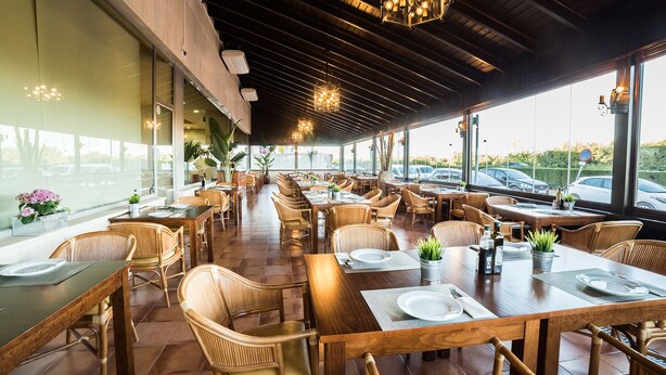 Restaurante com mesas e cadeiras de madeira, janelas amplas e vista para carros estacionados do lado de fora.