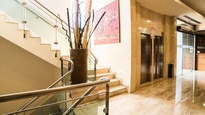 Um hall de hotel moderno com uma escadaria de vidro, um pilar de madeira com detalhes decorativos e uma pintura abstrata vermelha na parede.