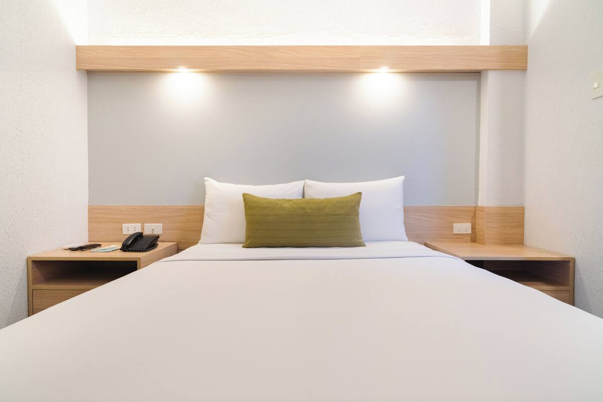 Quarto de hotel com uma cama, duas mesas de cabeceira, um telefone e uma luminária fixada na parede.