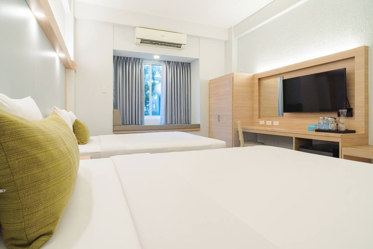 Quarto de hotel com duas camas, televisão, escrivaninha e janela com cortinas.