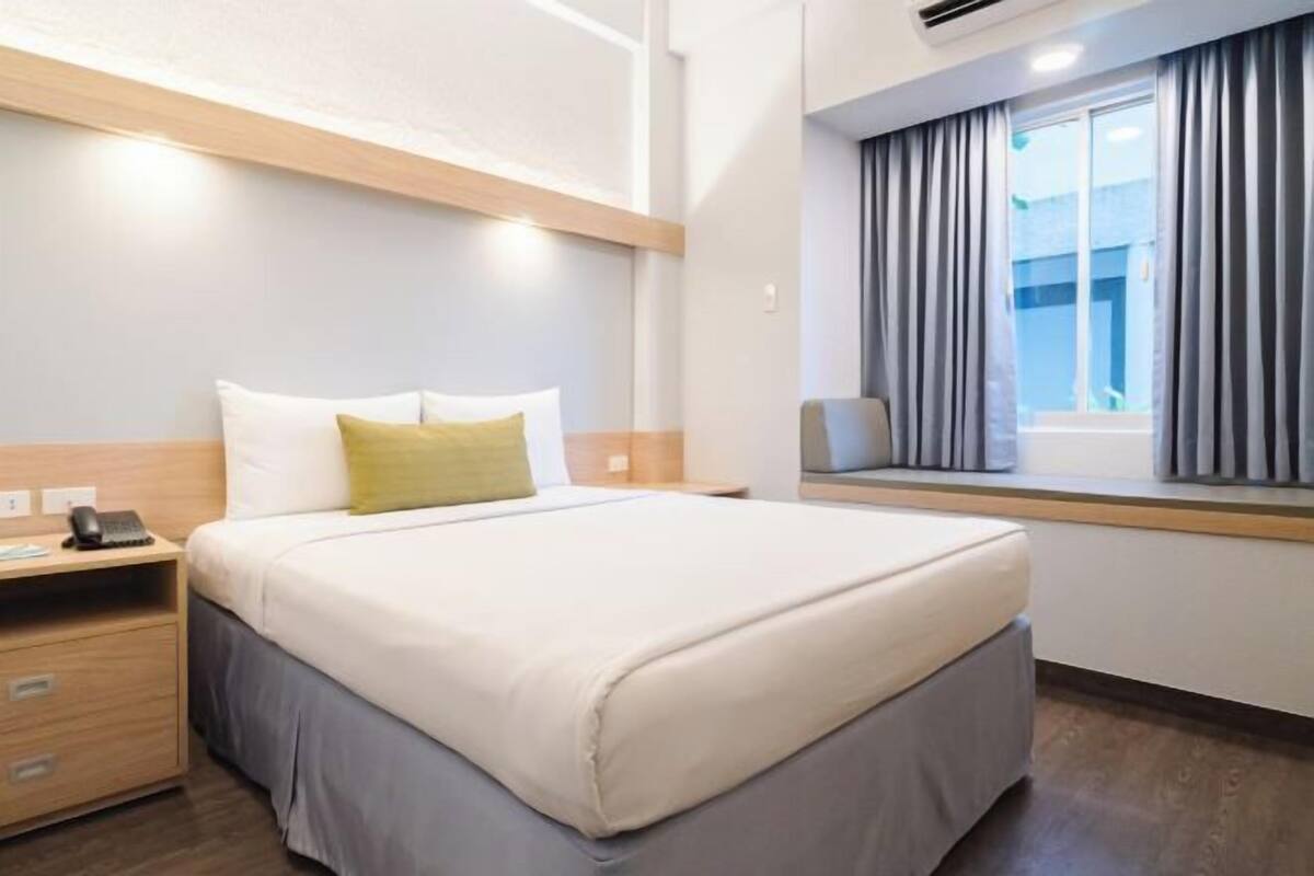 Quarto de hotel com cama, mesa de cabeceira e janela com cortinas.