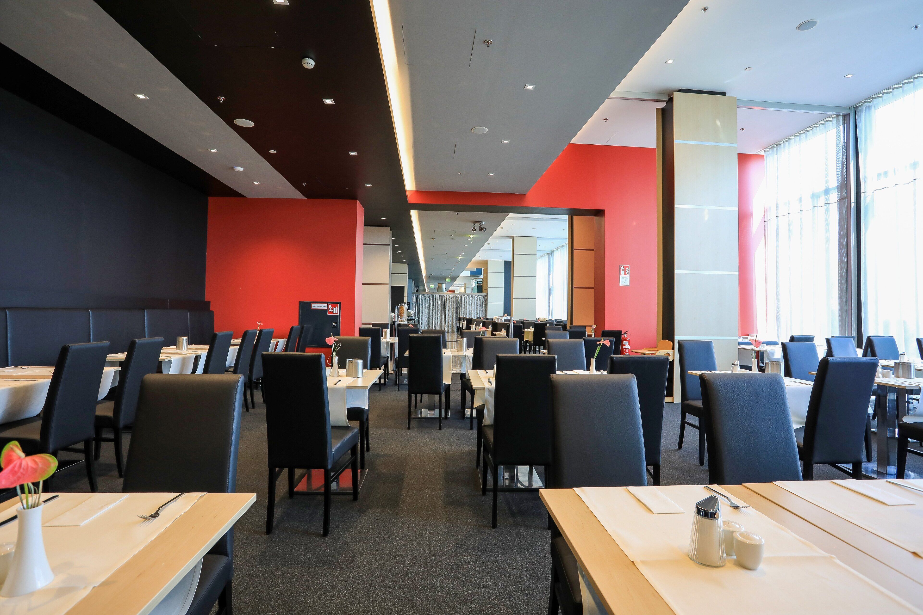 Uma sala de jantar moderna com várias mesas e cadeiras, uma parede de destaque vermelha e grandes janelas que permitem entrada de luz natural.