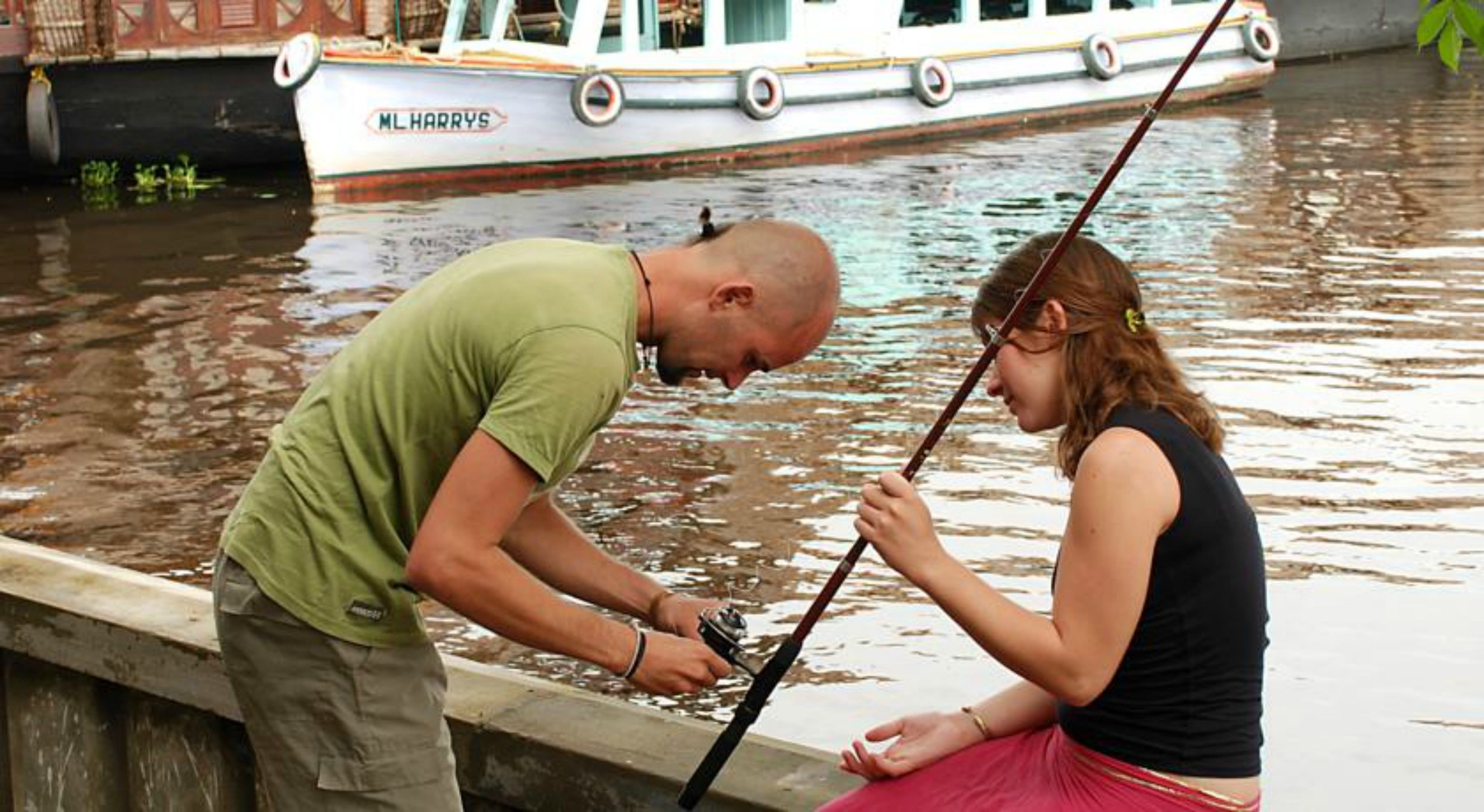 Duas pessoas pescam perto de um rio, com um barco chamado "ML HARRYS" ao fundo.
