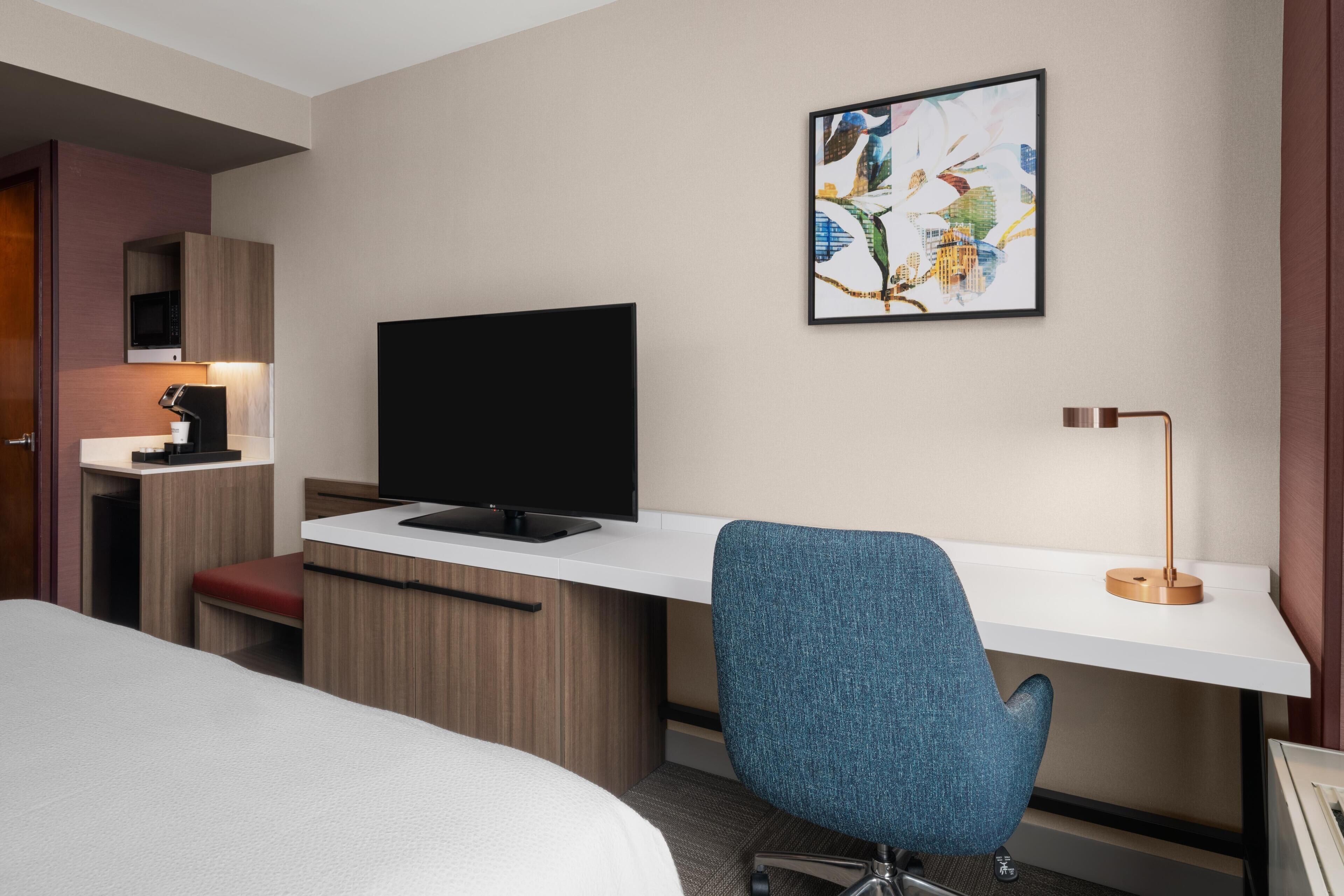 Quarto de hotel com cama, escrivaninha com televisão, cadeira, luminária de mesa, micro-ondas e cafeteira.