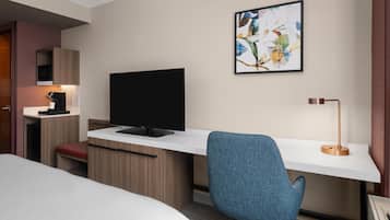 Quarto de hotel com cama, escrivaninha com televisão, cadeira, luminária de mesa, micro-ondas e cafeteira.
