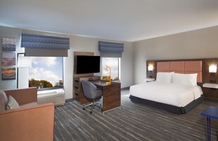 Quarto de hotel com cama, escrivaninha, cadeira, televisão e amplas janelas.